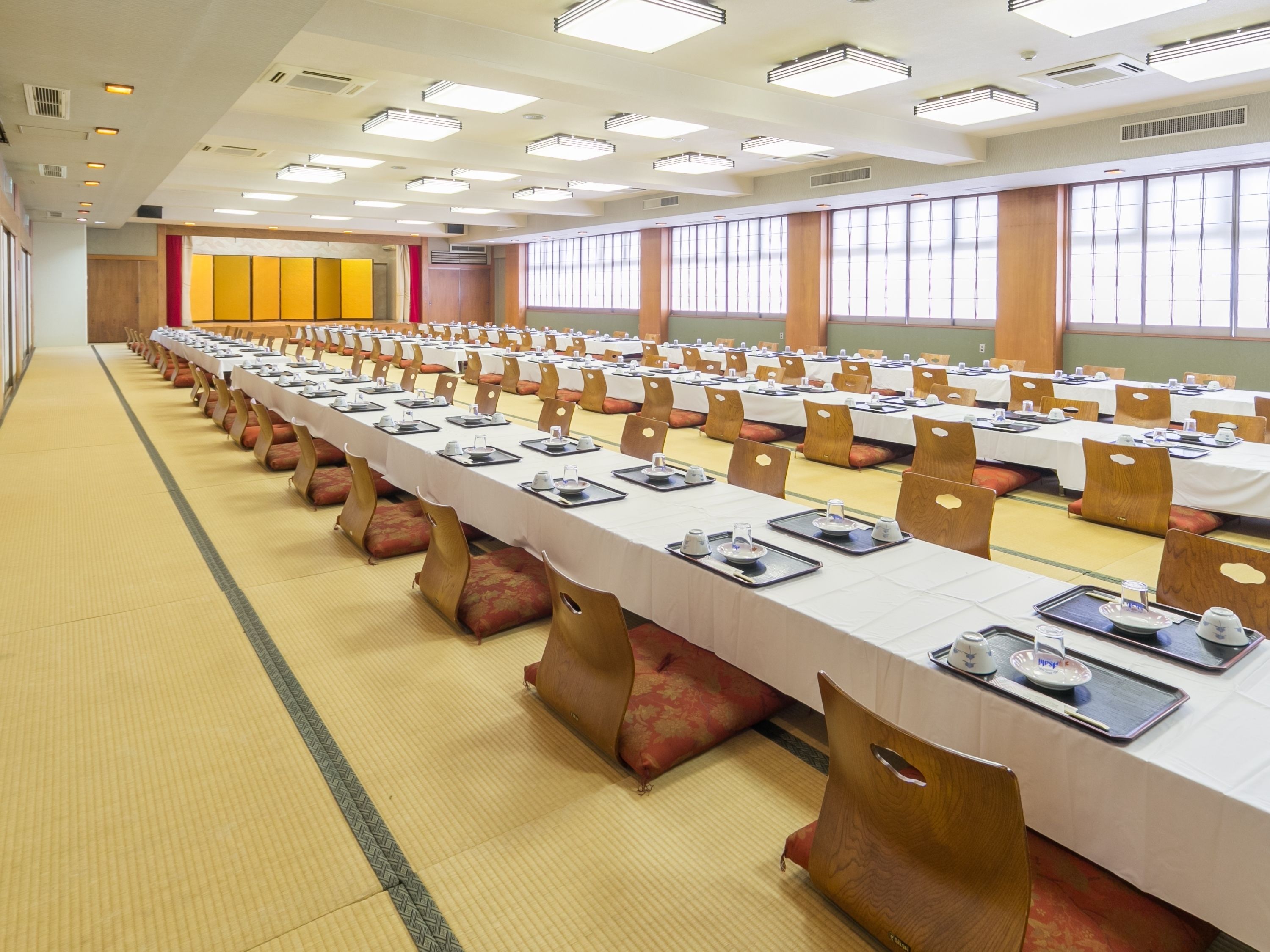 Sala de conferências com mesas compridas, cadeiras e teto alto com iluminação.
