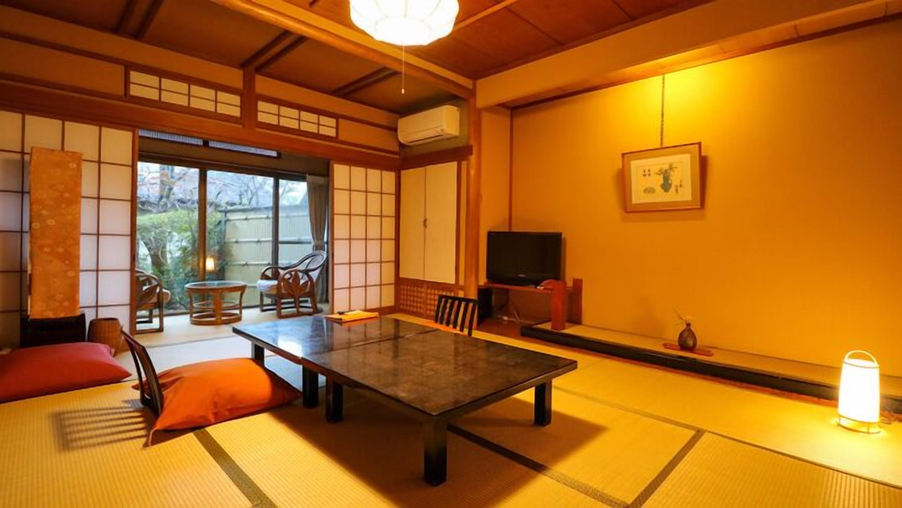 Um quarto japonês tradicional com tatames, uma mesa baixa, cadeiras, uma televisão e uma porta deslizante que dá para um jardim.