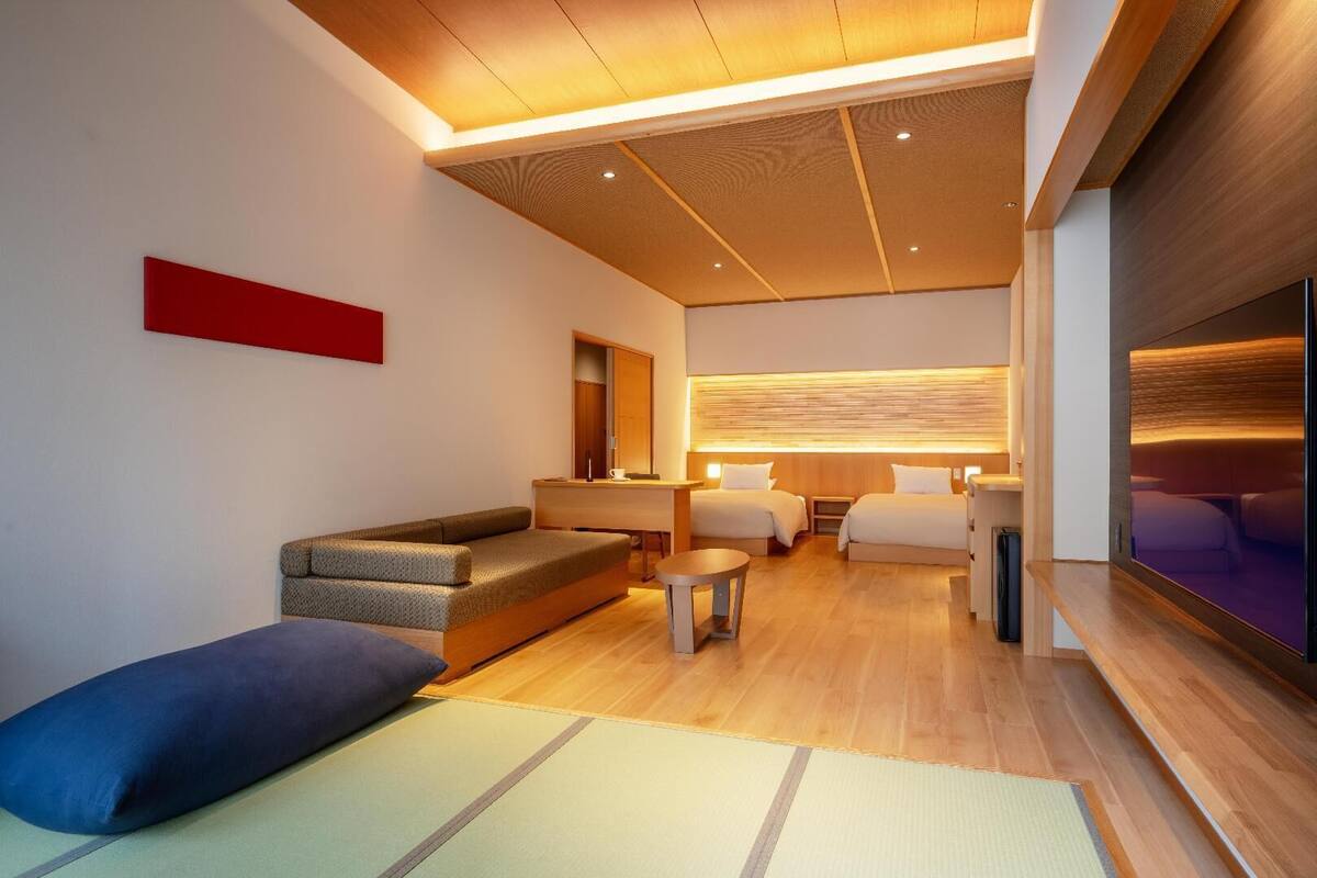 Quarto de hotel moderno com sofá, duas camas, uma mesinha e televisão.
