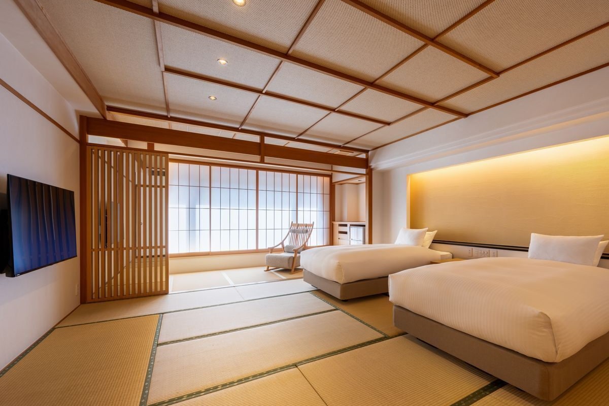Quarto de hotel tradicional em estilo japonês, com piso de tatame, teto baixo com vigas de madeira, porta deslizante e cama com roupa de cama branca.