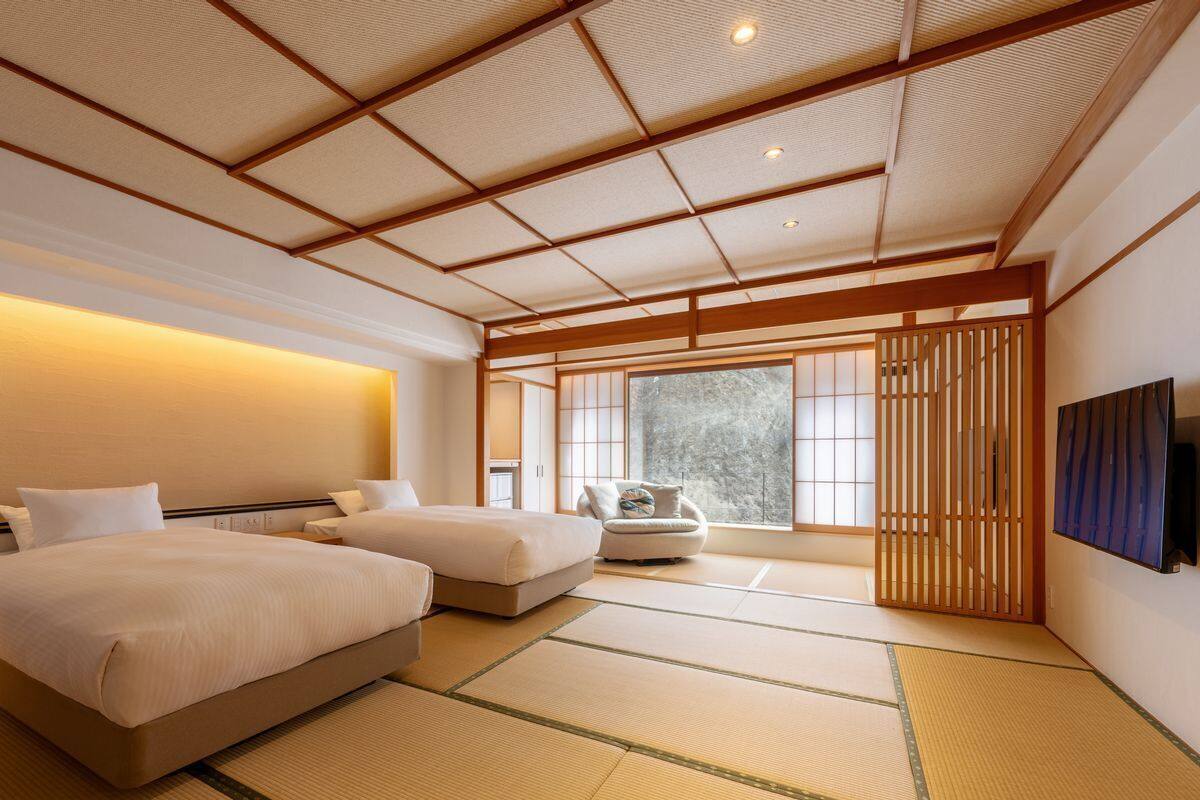 Quarto em estilo japonês tradicional, com tatames, uma cama, um sofá e uma porta deslizante para uma varanda.