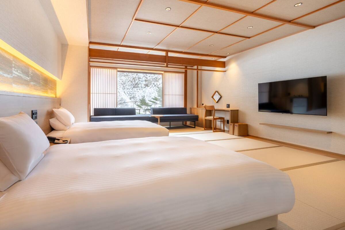 Quarto de hotel moderno com duas camas, televisão, escrivaninha e vista para árvores pela janela.