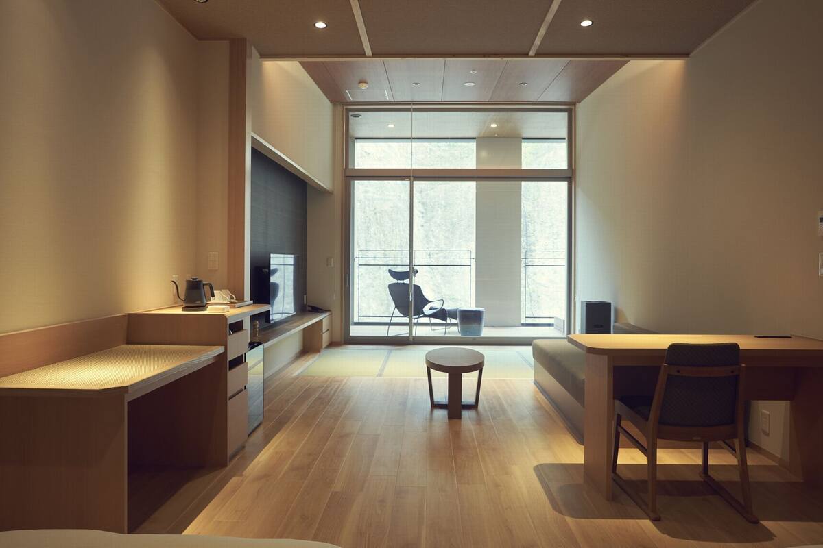 Quarto moderno e minimalista com escrivaninha de madeira, uma cadeira, uma mesinha e um sofá. Há uma janela grande que oferece luz natural e vista para o exterior.