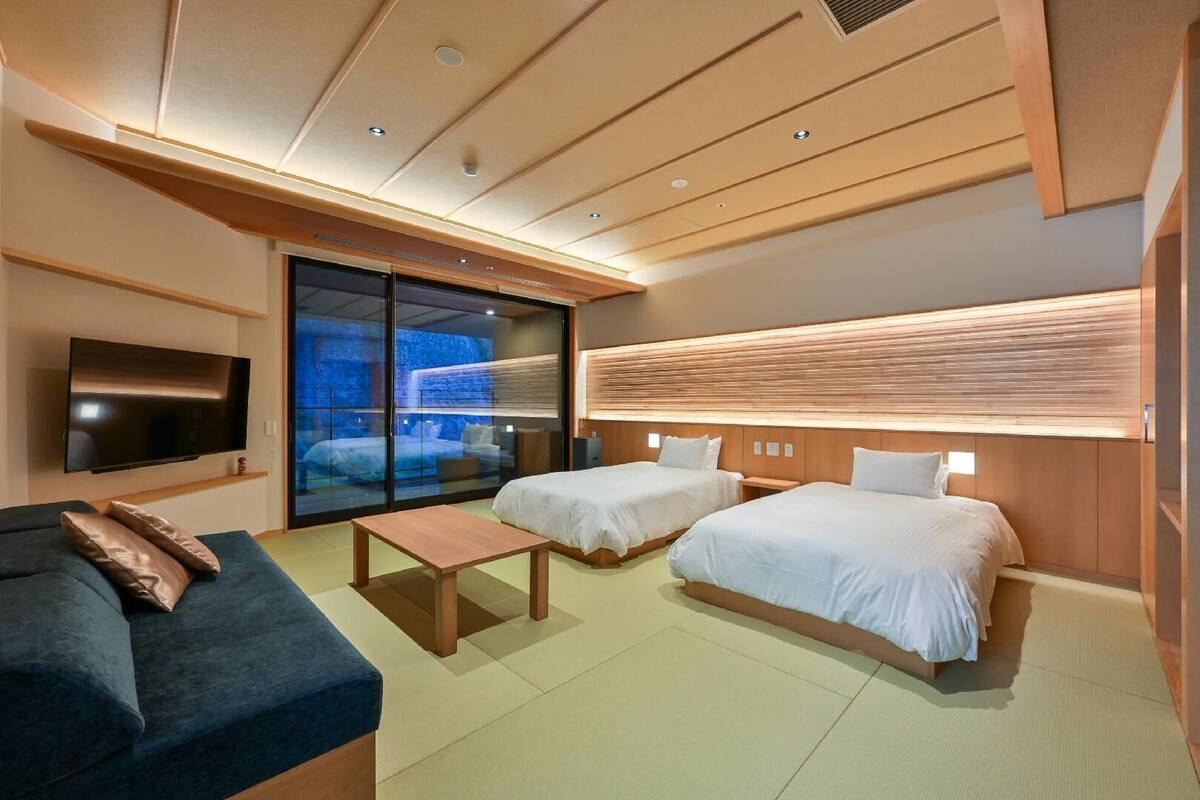 Quarto de hotel moderno com televisão de tela plana, duas camas, um sofá, porta de vidro deslizante e teto de madeira.