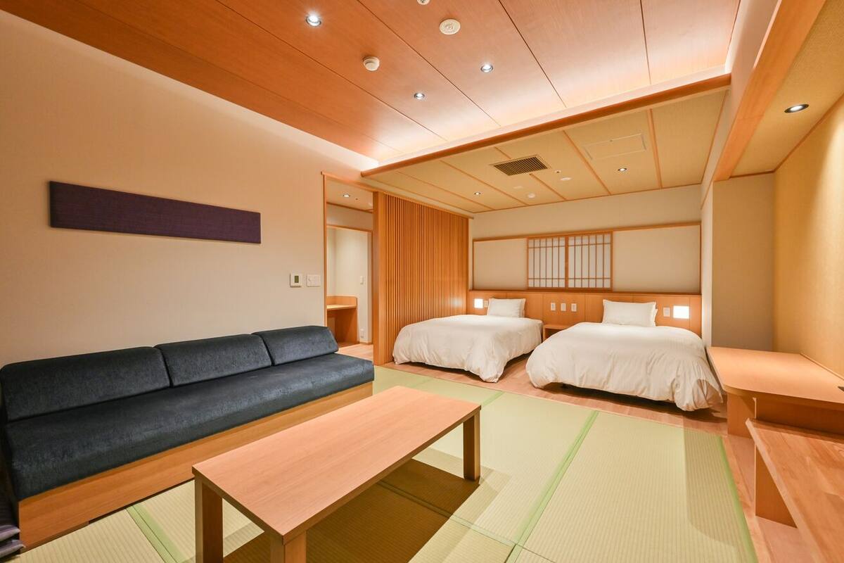 Quarto de hotel com sofá, duas camas, uma mesa de madeira, esteiras de tatame e uma porta japonesa tradicional deslizante.