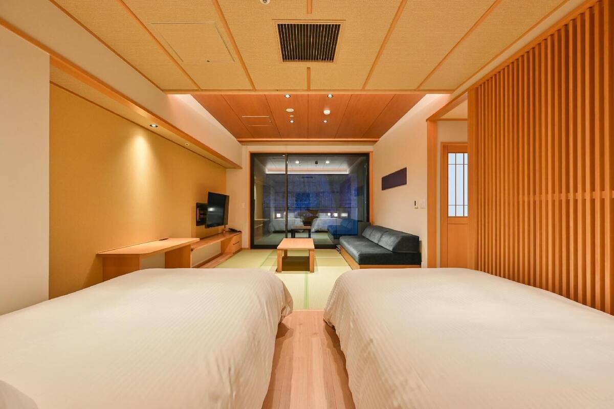 Quarto de hotel com duas camas, televisão, sofá e porta de vidro deslizante para uma varanda.
