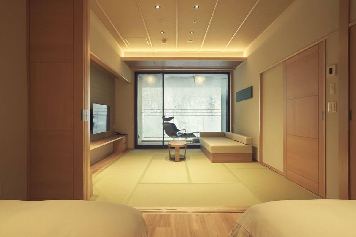 Quarto de hotel moderno com porta de vidro deslizante, um sofá, uma mesinha e uma televisão.