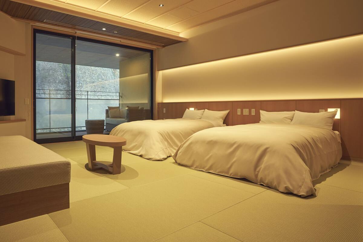 Quarto de hotel com duas camas, um sofá, uma mesinha, uma televisão e uma porta de correr para o exterior.