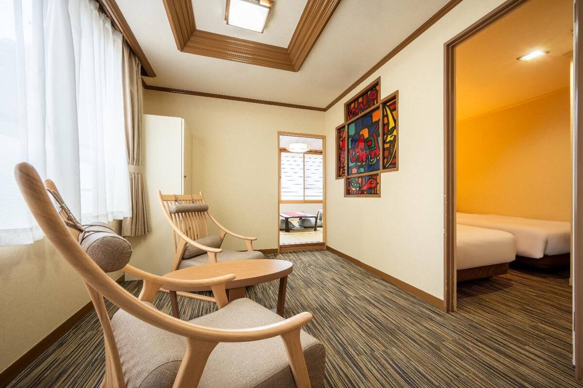 Quarto de hotel com cama, uma cadeira, mesa, janela com cortinas e parede com arte colorida.