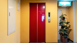 Um corredor com elevador vermelho, sinal de saída de emergência fixado na parede e um vaso de planta.