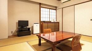Um quarto japonês tradicional com tatames, uma mesa de madeira, cadeiras, uma televisão, um frigorífico e portas deslizantes.