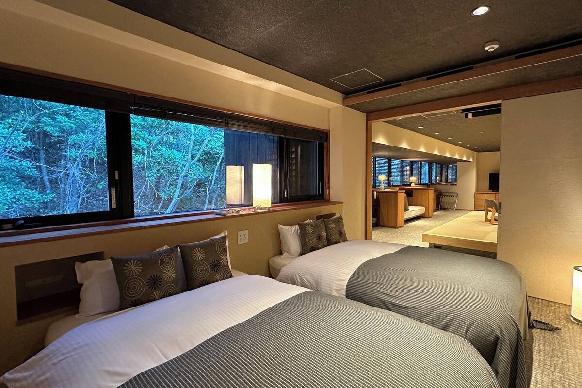 Quarto de hotel com duas camas, uma janela grande com vista para árvores, uma escrivaninha com cadeira e um pequeno espaço de estar.