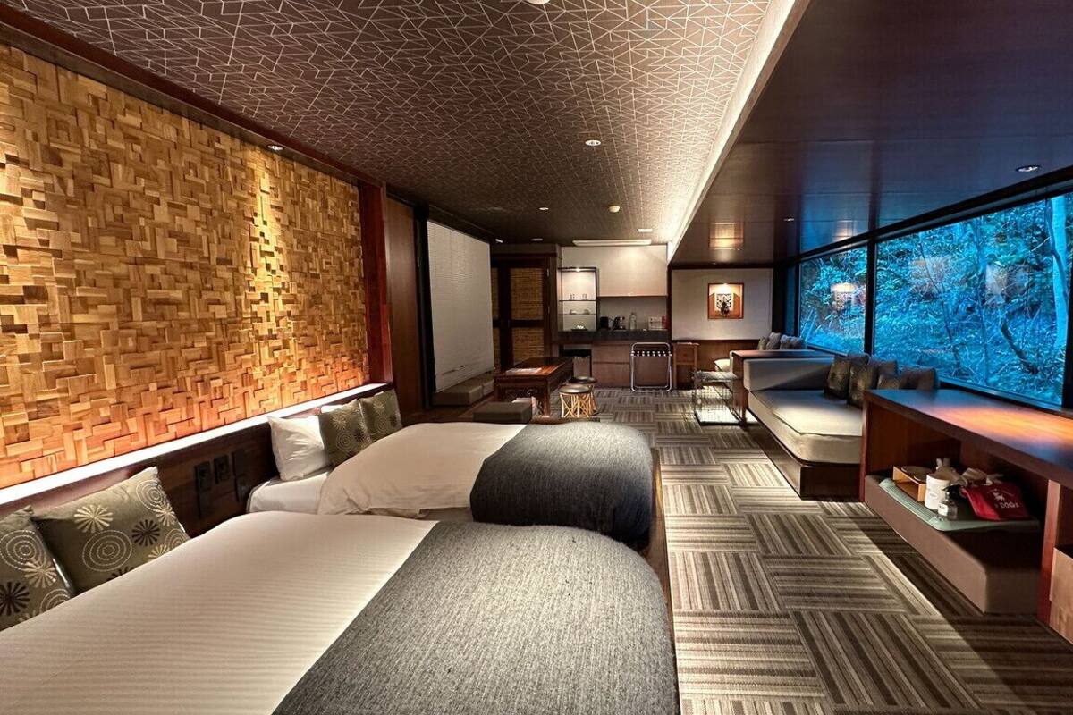 Quarto de hotel com uma janela grande, duas camas, um espaço de estar e vista para uma floresta.