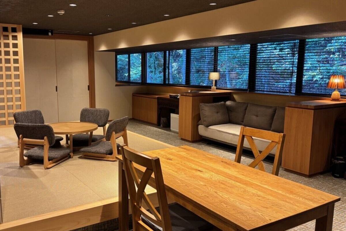 Interior moderno com mesa de jantar de madeira, cadeiras estofadas em cinza, um sofá e uma janela ampla com vista para árvores.