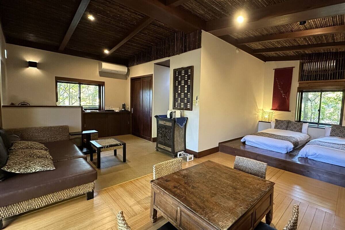 Quarto com sofá, mesa de madeira, duas camas com cabeceiras, um objeto decorativo na parede e janela com vista.