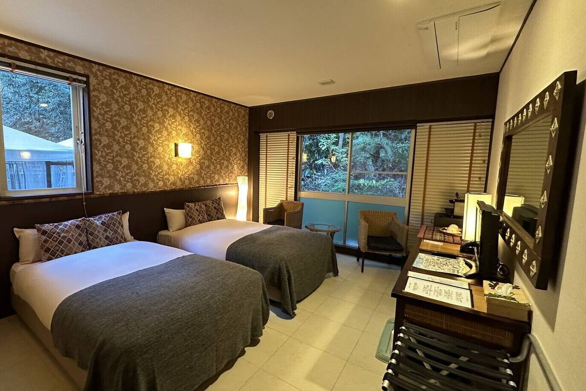 Quarto de hotel com duas camas, uma escrivaninha com computador e uma janela grande com cortinas.
