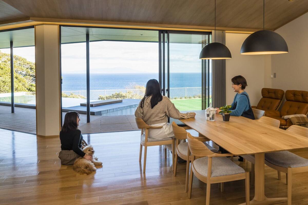 Interior moderno com mesa de jantar de madeira, duas cadeiras, um sofá e uma janela grande com vista para o mar.