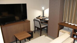 Quarto de hotel compacto com televisão de tela plana, escrivaninha com luminária, cadeira, mesa pequena e sofá.