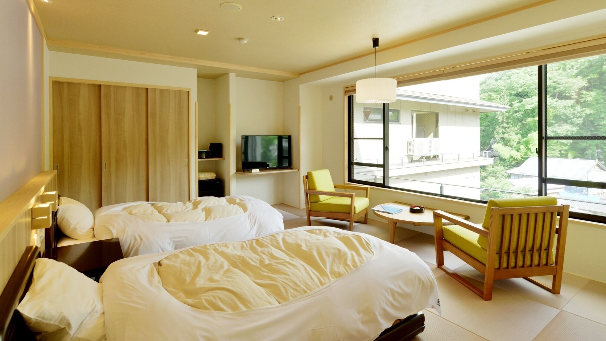 Quarto de hotel com duas camas, uma televisão, uma cadeira e uma janela grande com vista para edifícios e árvores.