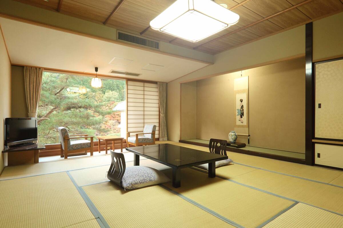 Um quarto japonês tradicional com tatames, uma mesa baixa, cadeiras, uma televisão e vista para árvores.