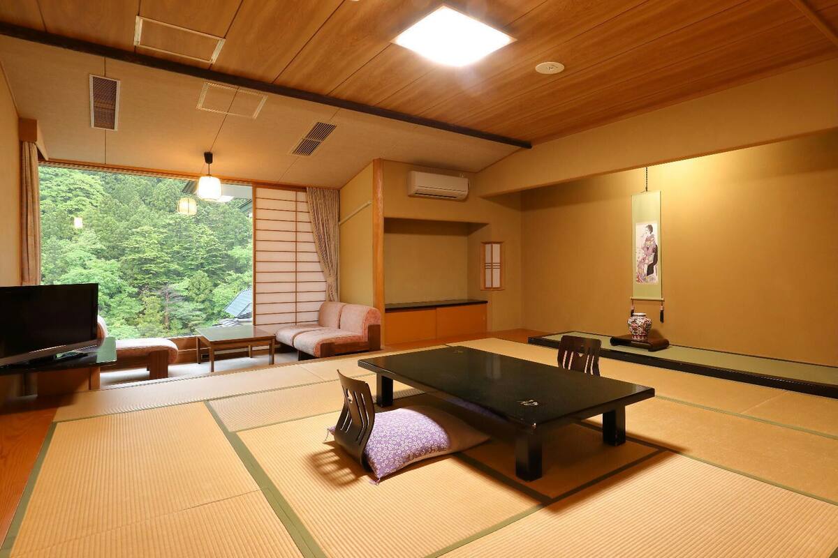 Um espaço com tatames, incluindo uma mesa de tatame, um sofá, uma televisão e uma porta deslizante que dá para um visual com vegetação.