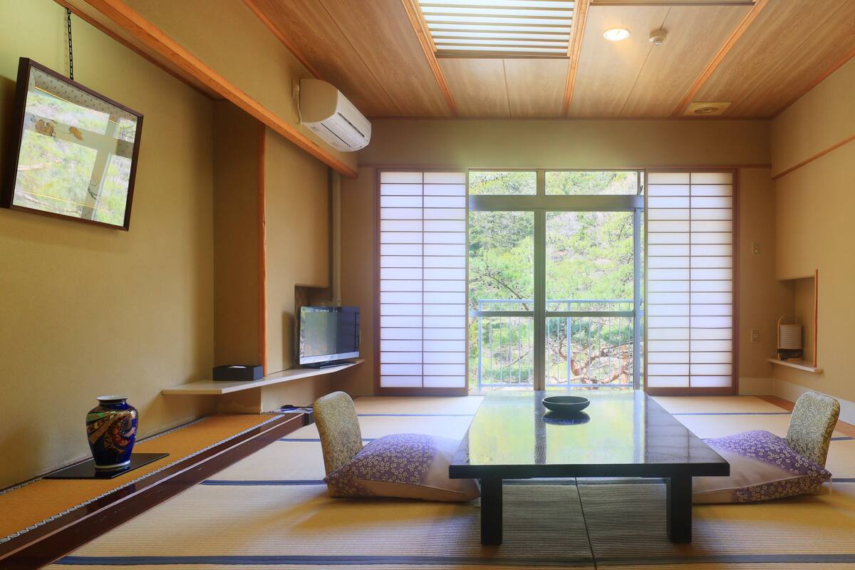 Um quarto japonês tradicional com tatames, uma mesa baixa, dois assentos de piso e uma televisão, além de uma grande janela com portas deslizantes.