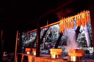 Uma cozinha tradicional com milho pendurado e panelas a fumegar, em um espaço ao ar livre sombreado e com vista para árvores e vegetação.