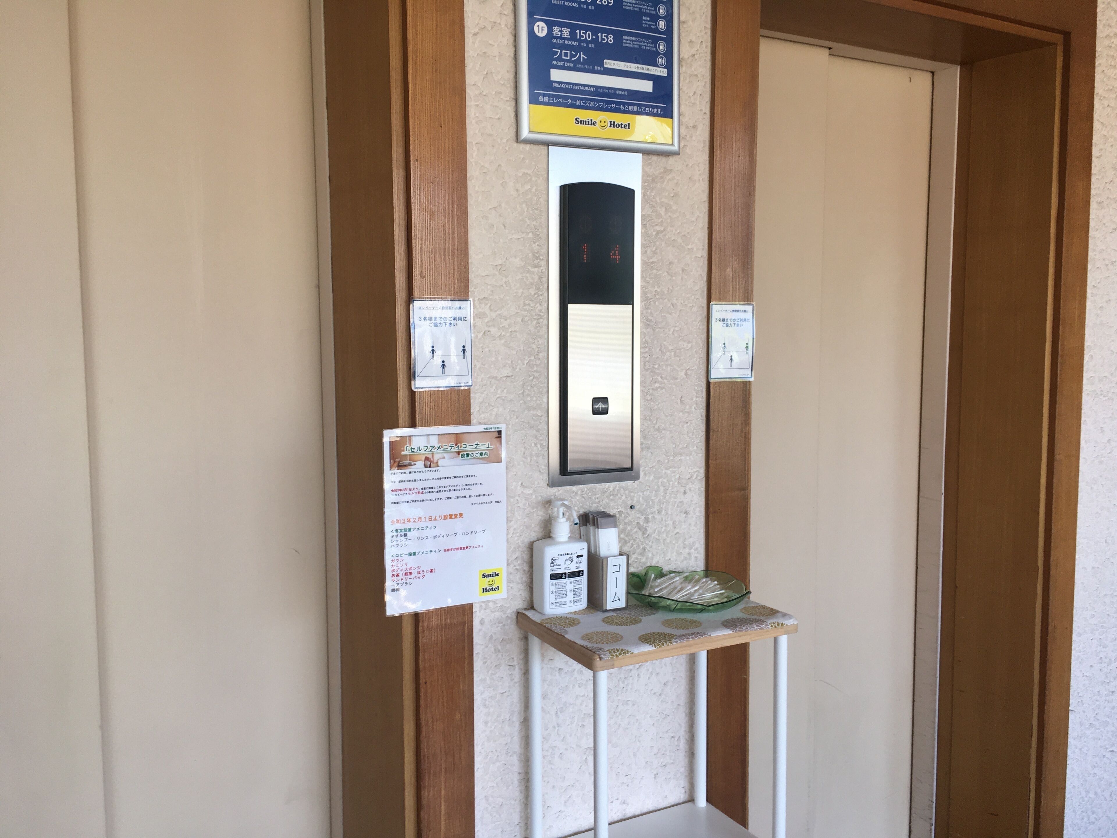 Um elevador com uma placa indicando o primeiro andar. Há placas informativas e uma pequena mesa com itens como álcool em gel e uma tigela de vidro.
