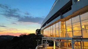 Um edifício moderno com amplas janelas de vidro que refletem luzes internas, cercado por vegetação.