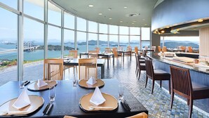 Restaurante com mesas preparadas para refeições, grandes janelas com vista para uma ponte e colinas, e piso de azulejos.