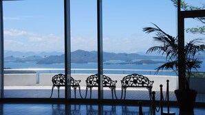 Vista através de grandes janelas de uma varanda com três cadeiras de metal ornamentadas e um vaso de plantas, com vista para um corpo de água e montanhas ao longe.