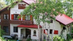 Uma casa de dois andares com telhado vermelho e paredes brancas, rodeada por vegetação.