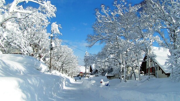 Rua coberta de neve com casas em ambos os lados, céu azul límpido e uma pessoa vestida de boneco de neve.