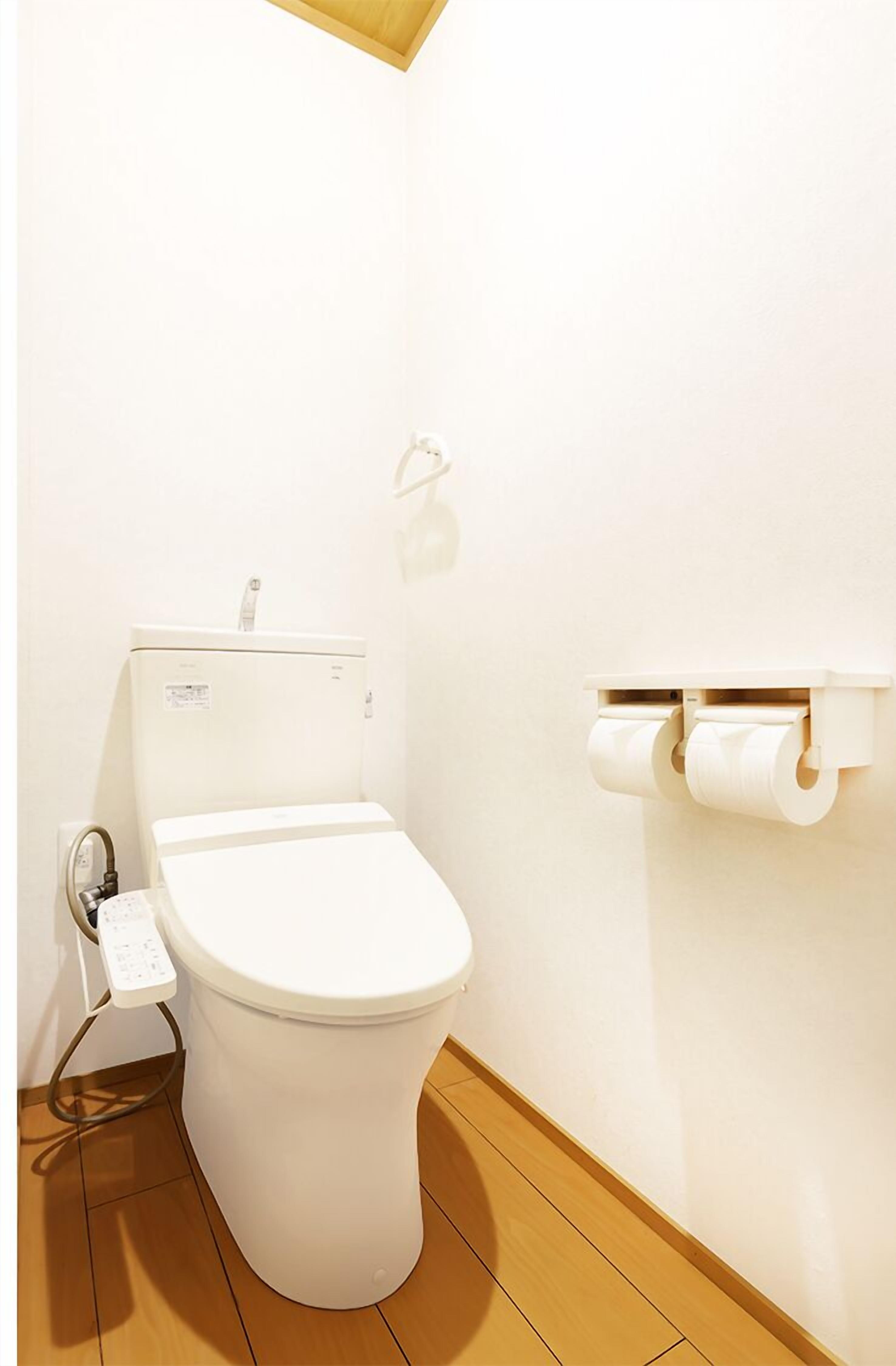 Sanitário branco com painel de controle, rolo de papel higiênico na parede e chão de madeira.