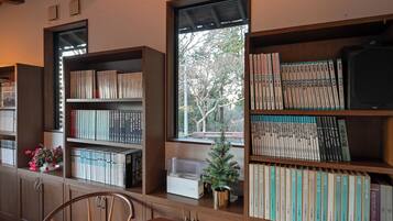Uma biblioteca com estantes cheias de livros, um pequeno pinheiro de Natal e uma janela com vista para árvores.