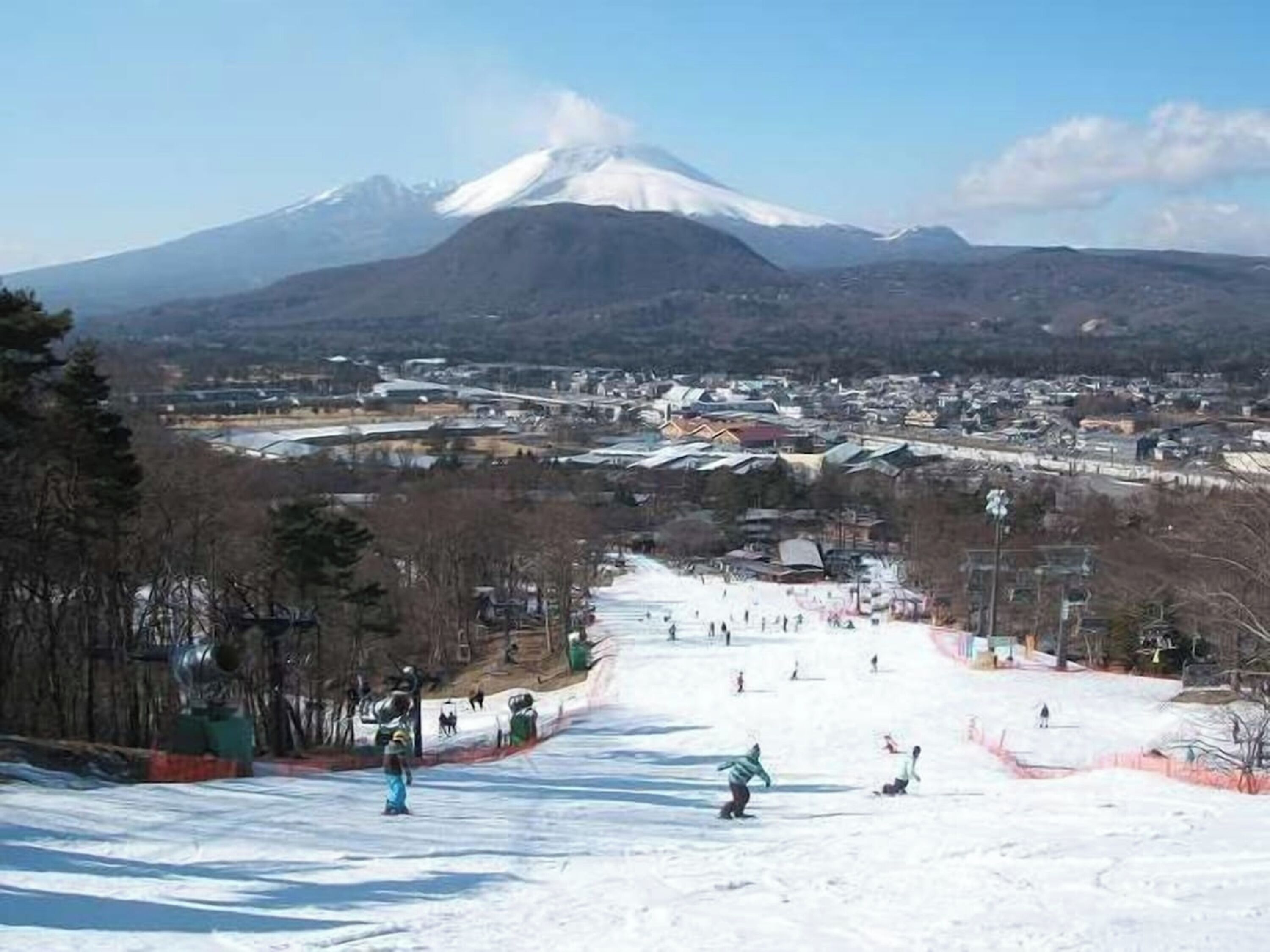 Pista de esqui com esquiadores, uma cidade ao fundo e uma montanha coberta de neve.
