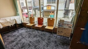 Quarto com piso carpetado, três cadeiras de madeira, janela com vista para edifícios e cortina à esquerda.