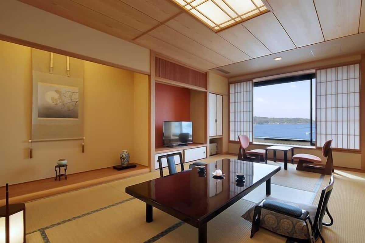 Um quarto tradicional japonês com tatames, uma mesa baixa, cadeiras, uma televisão e uma grande janela com vista para o mar.