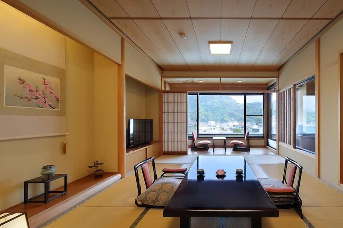 Um quarto tradicional japonês com piso de tatame, uma mesa baixa com almofadas, uma televisão e vista para uma paisagem montanhosa.