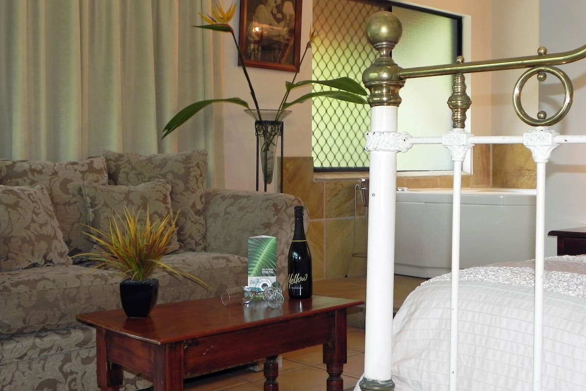 Quarto com cama branca, mesa de centro de madeira com uma planta, sofá e banheiro visível por uma janela.