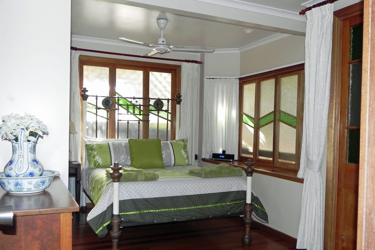 Quarto com cama, mesa de cabeceira, vaso com flores, ventoinha no teto e janela com cortinas.