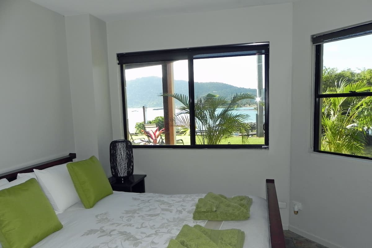 Quarto com cama, almofadas verdes, mesa de cabeceira e janela com vista para um corpo de água e vegetação.