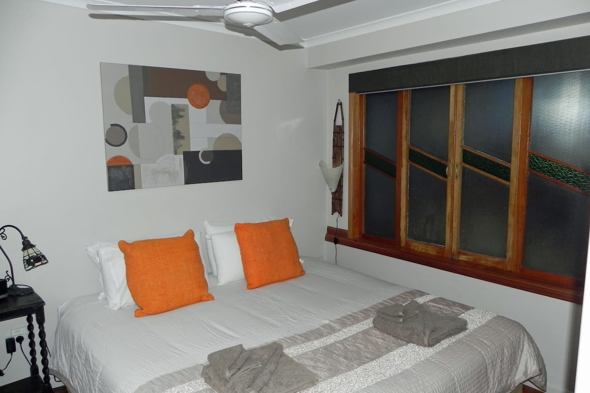 Quarto com cama, duas almofadas laranja, uma almofada branca, uma toalha cinza, cabeceira de madeira, janela com vista, ventilador de teto e uma obra de arte na parede.