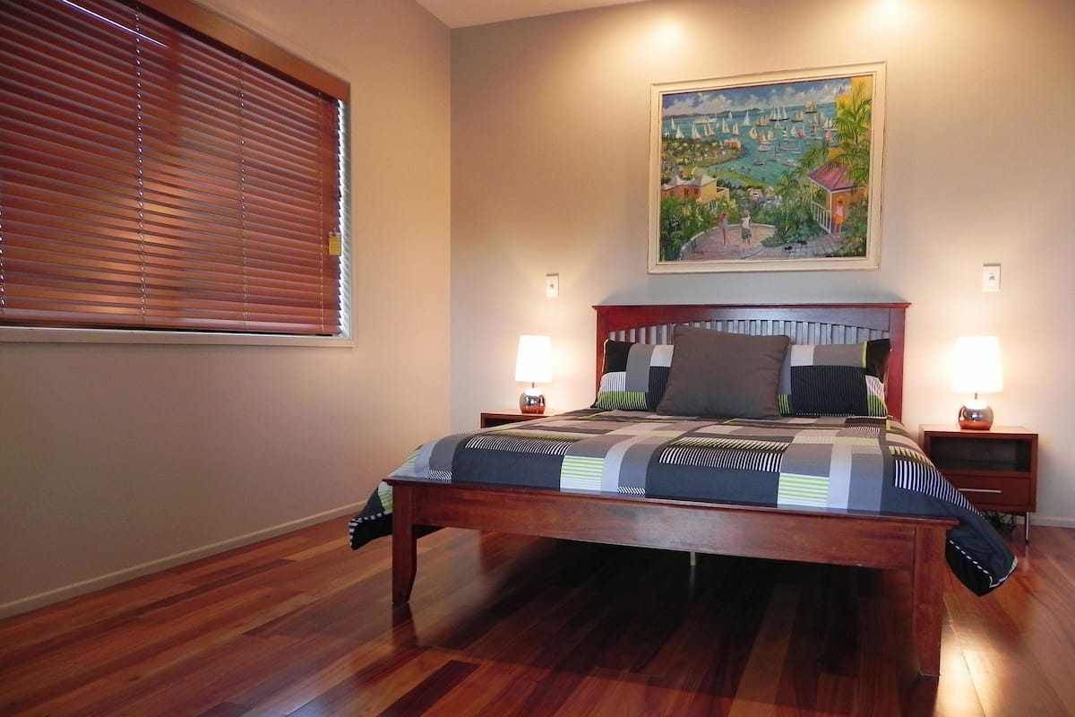 Um quarto com cama de madeira, duas mesas de cabeceira com abajures, um quadro na parede e uma janela com persianas.