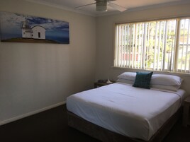 Quarto com cama, mesa de cabeceira, luminária, quadro de farol e janela com persianas.