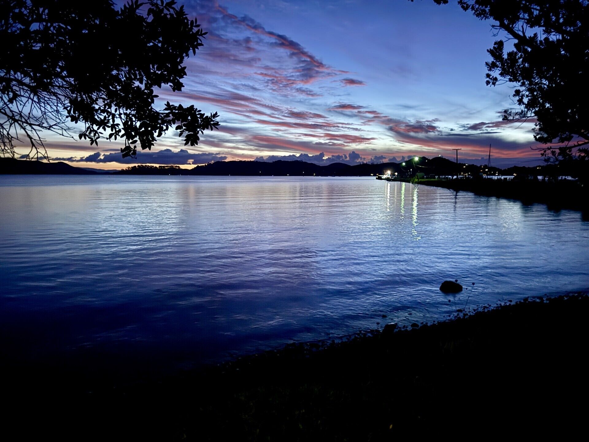 Um lago tranquilo ao anoitecer com céu em tons de pôr do sol, árvores emoldurando a paisagem e uma linha de costa distante com luzes.