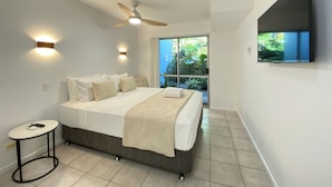 Quarto moderno com cama, ventoinha no teto, TV na parede e vista para o verde lá fora.