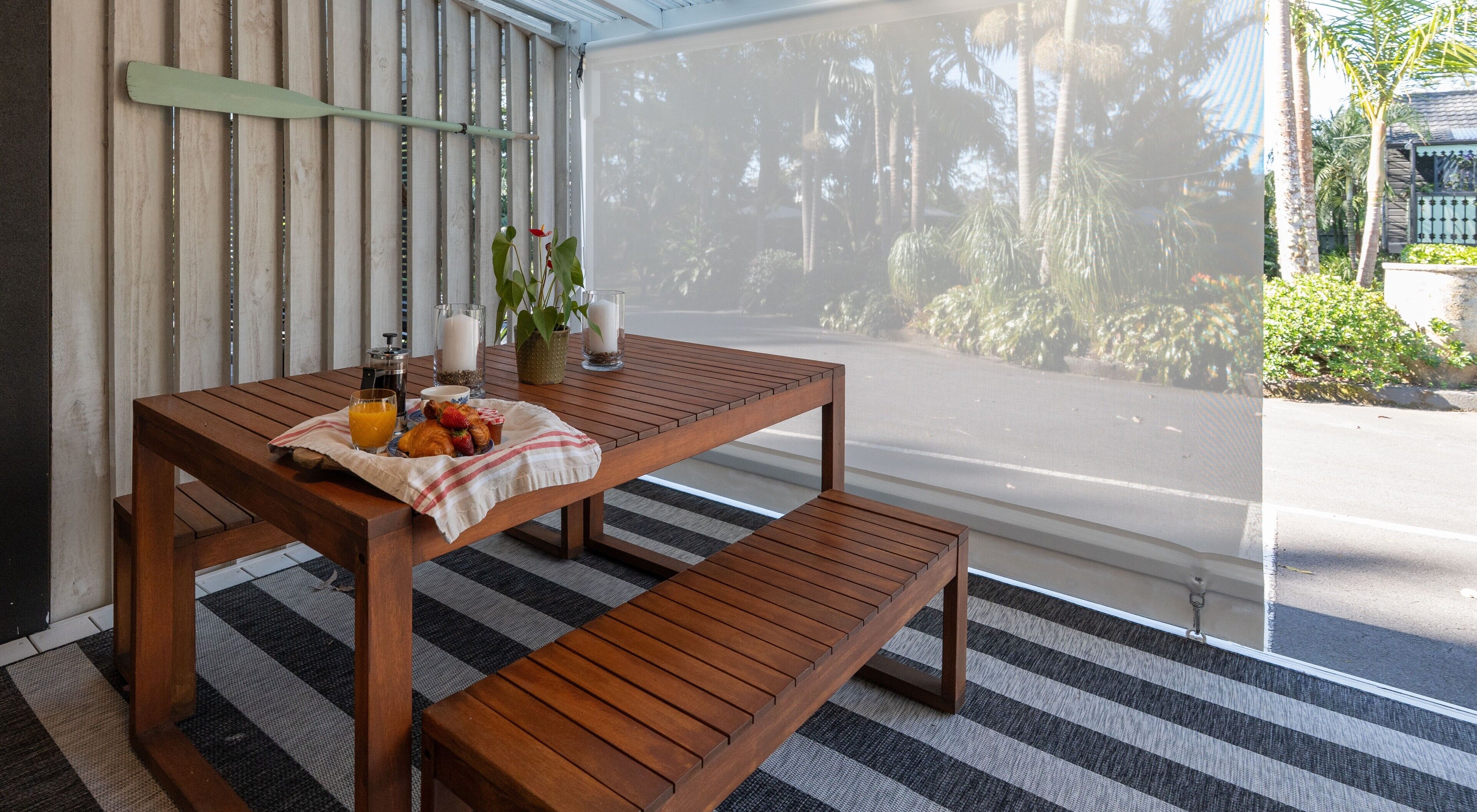 Uma mesa de madeira com toalha xadrez, um banco e uma disposição para café da manhã com croissants, um copo de suco de laranja e uma vela.