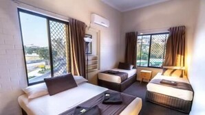 Quarto de hotel com duas camas, uma mesinha, uma mesa de cabeceira e janela com cortinas.