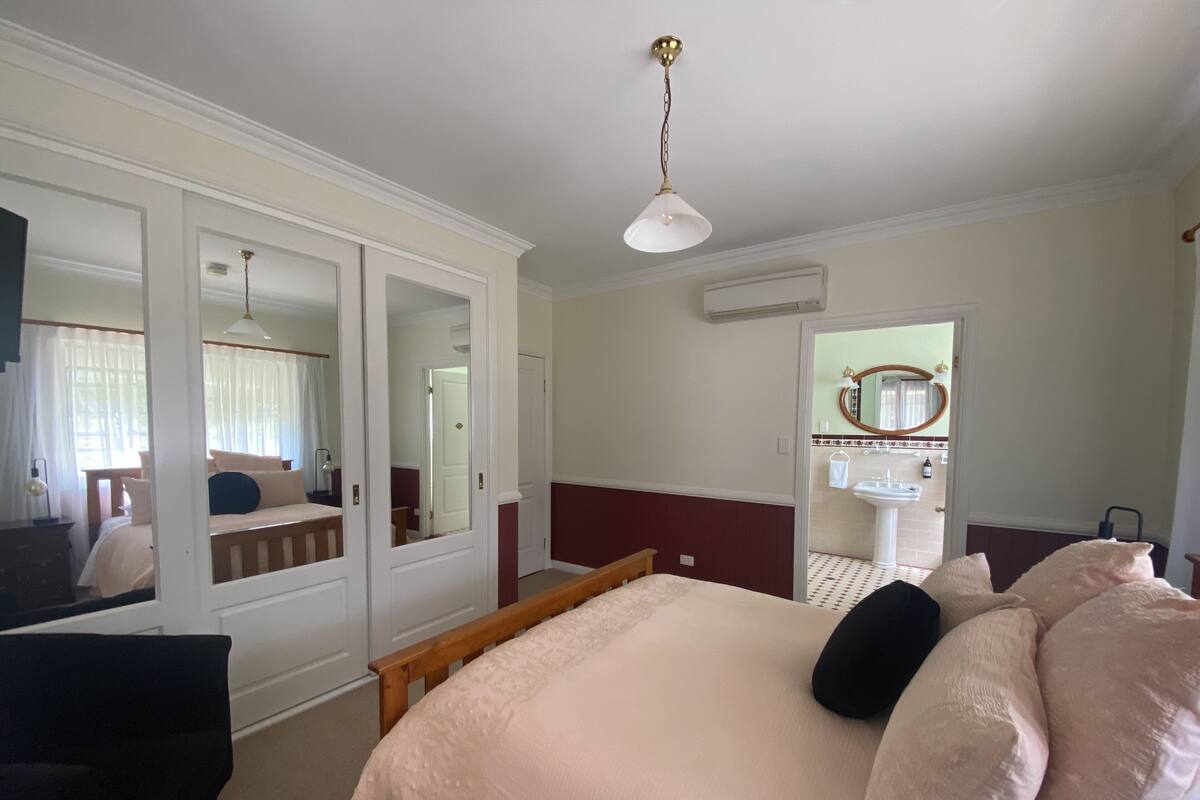 Quarto com cama, um banquinho de madeira, um espelho e banheiro com pia e espelho.